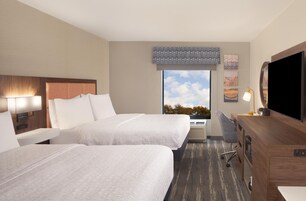 Quarto de hotel com duas camas, uma escrivaninha, televisão e janela com vista para árvores.