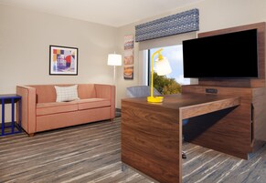Quarto de hotel com escrivaninha de madeira, televisão, sofá, luminária e quadro na parede.