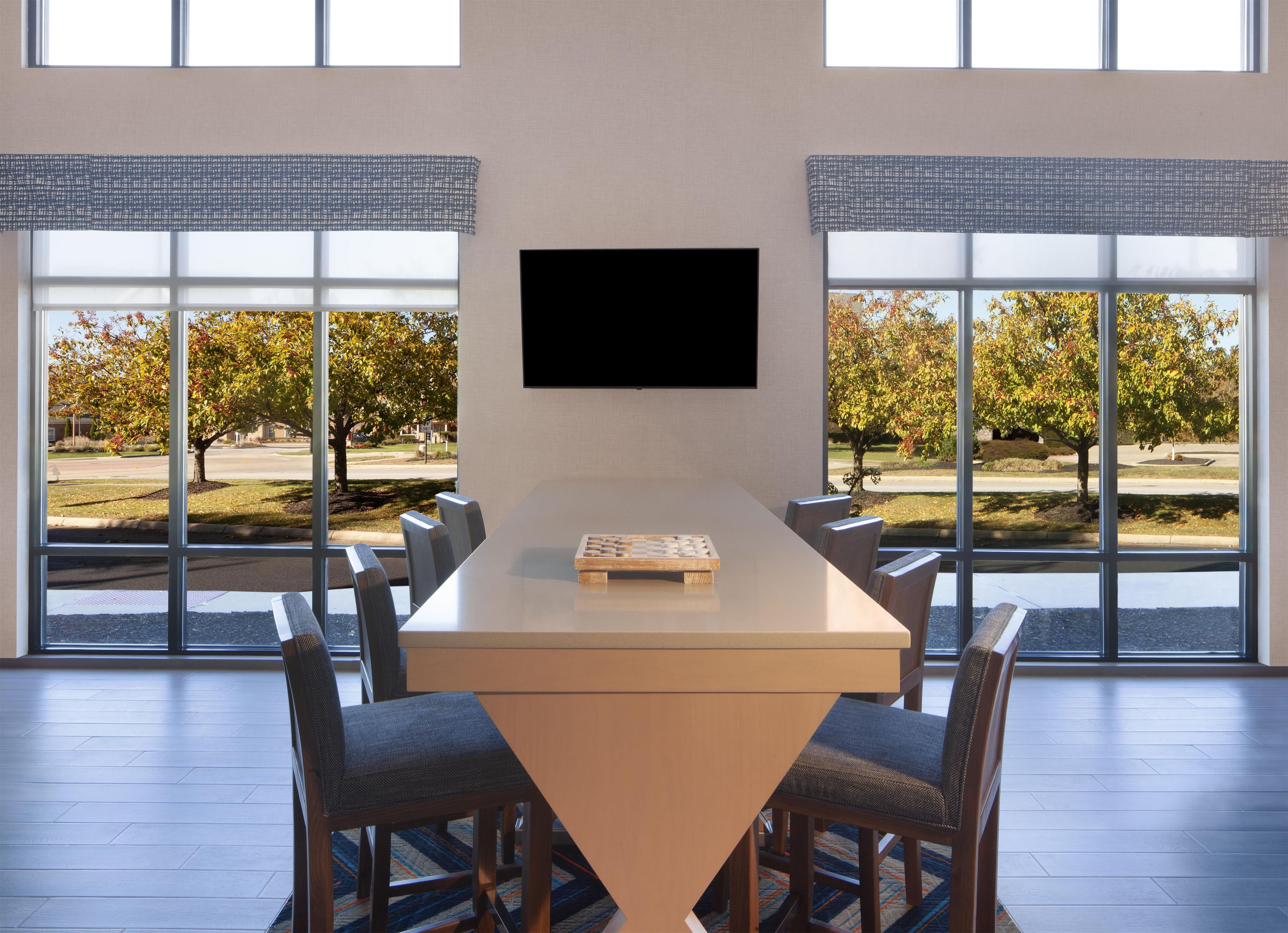 Sala de conferências com uma mesa grande, seis cadeiras, uma TV de tela plana e janelas amplas com vista para árvores e uma rua.