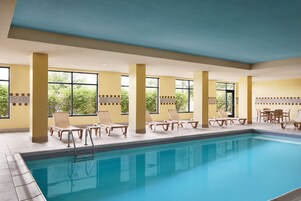 Piscina com espreguiçadeiras e uma mesa, cercada por janelas com vista para o verde.
