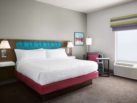 Quarto de hotel com uma cama grande, duas mesas de cabeceira, uma cadeira, janela com persianas e ar condicionado fixado na parede.