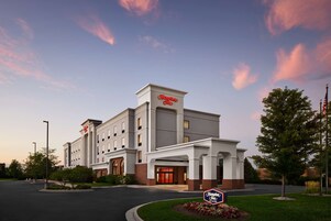 Hotel Hampton Inn com estilo arquitetônico clássico, apresentando entrada central com porta vermelha, cercada por um gramado bem cuidado e área de estacionamento.