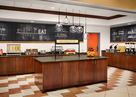 Um balcão de café da manhã com uma placa indicando o início do café, uma cesta de frutas e diversos alimentos no balcão.
