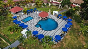 Piscina com espreguiçadeiras e guarda-sóis, cercada por palmeiras e um edifício com telhado vermelho.