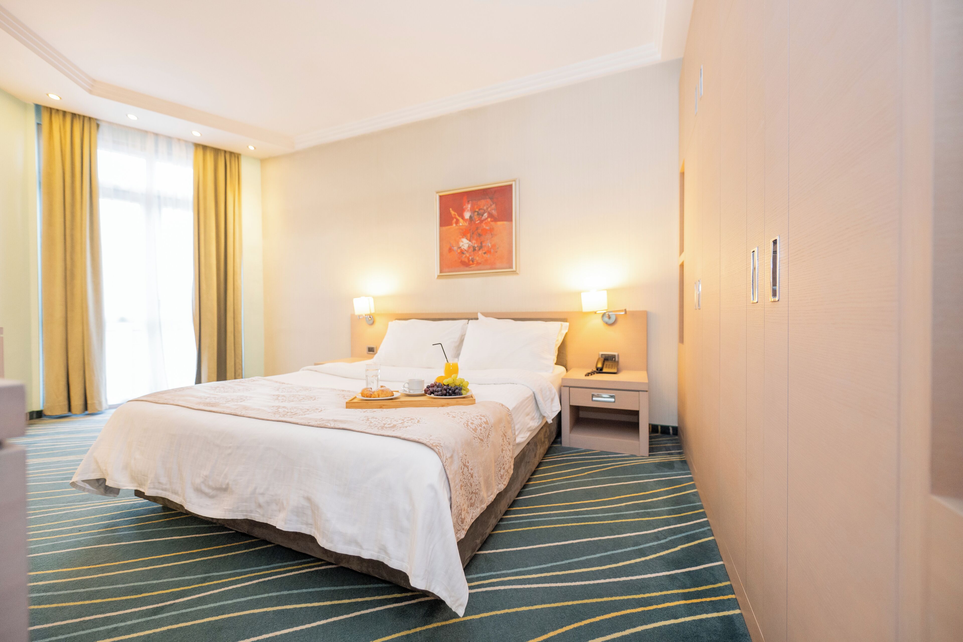 Quarto de hotel com uma cama grande, duas mesas de cabeceira, um criado-mudo, um quadro na parede e uma janela com cortinas.