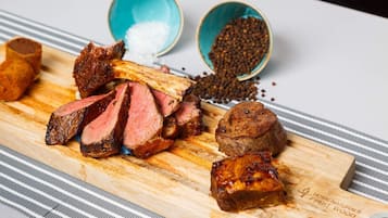 Tábua de madeira com carne fatiada, um osso de carne inteiro e duas pequenas tigelas com pimenta do reino e sal.