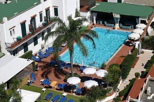 Piscina no terraço com espreguiçadeiras e guarda-sóis, cercada por um edifício com varandas e uma palmeira.