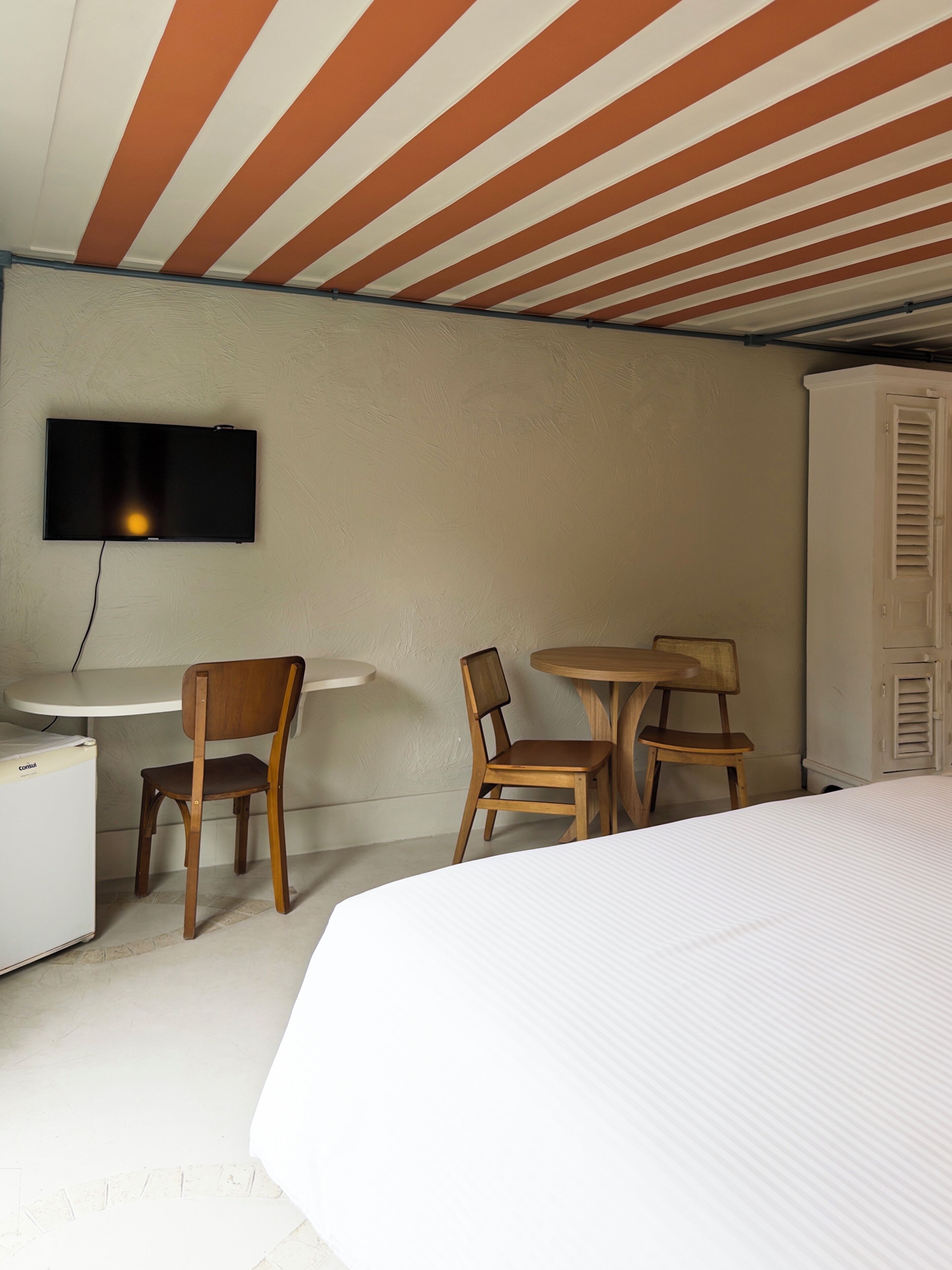 Quarto de hotel com cama, escrivaninha, duas cadeiras, televisão e guarda-roupa.