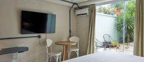 Quarto de hotel com uma cama, televisão fixada na parede, uma pequena mesa com cadeiras e uma porta de vidro deslizante para uma área externa com plantas.