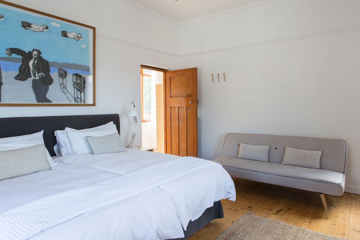 Quarto com cama, sofá, piso de madeira, porta e um quadro na parede.