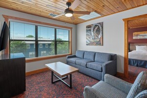 Quarto com ventilador de teto, uma janela grande com persianas, sofá cinza, mesa pequena e cama com cabeceira de madeira.