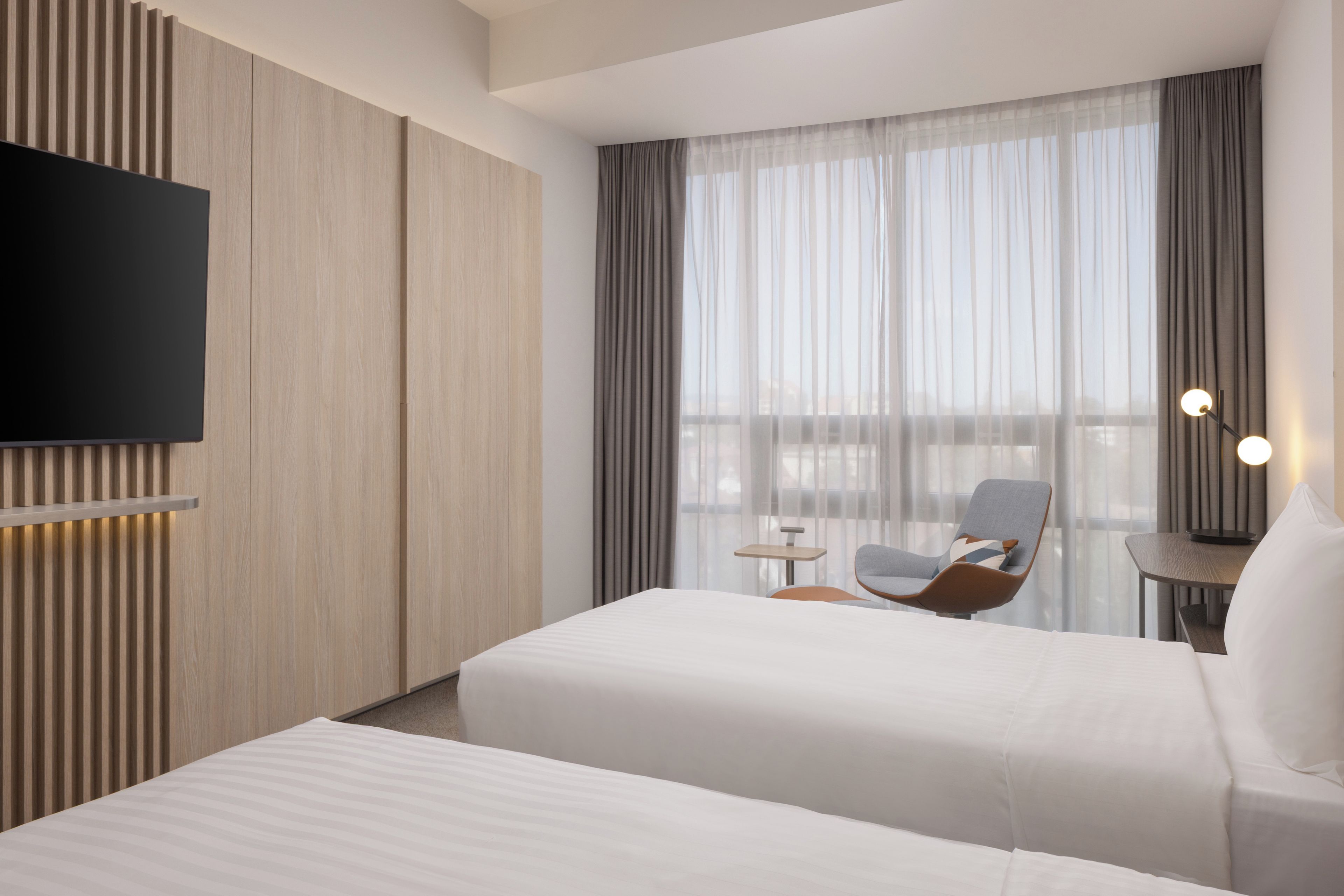 Quarto de hotel com duas camas, televisão de tela plana, uma mesinha, uma cadeira e janela com cortinas.