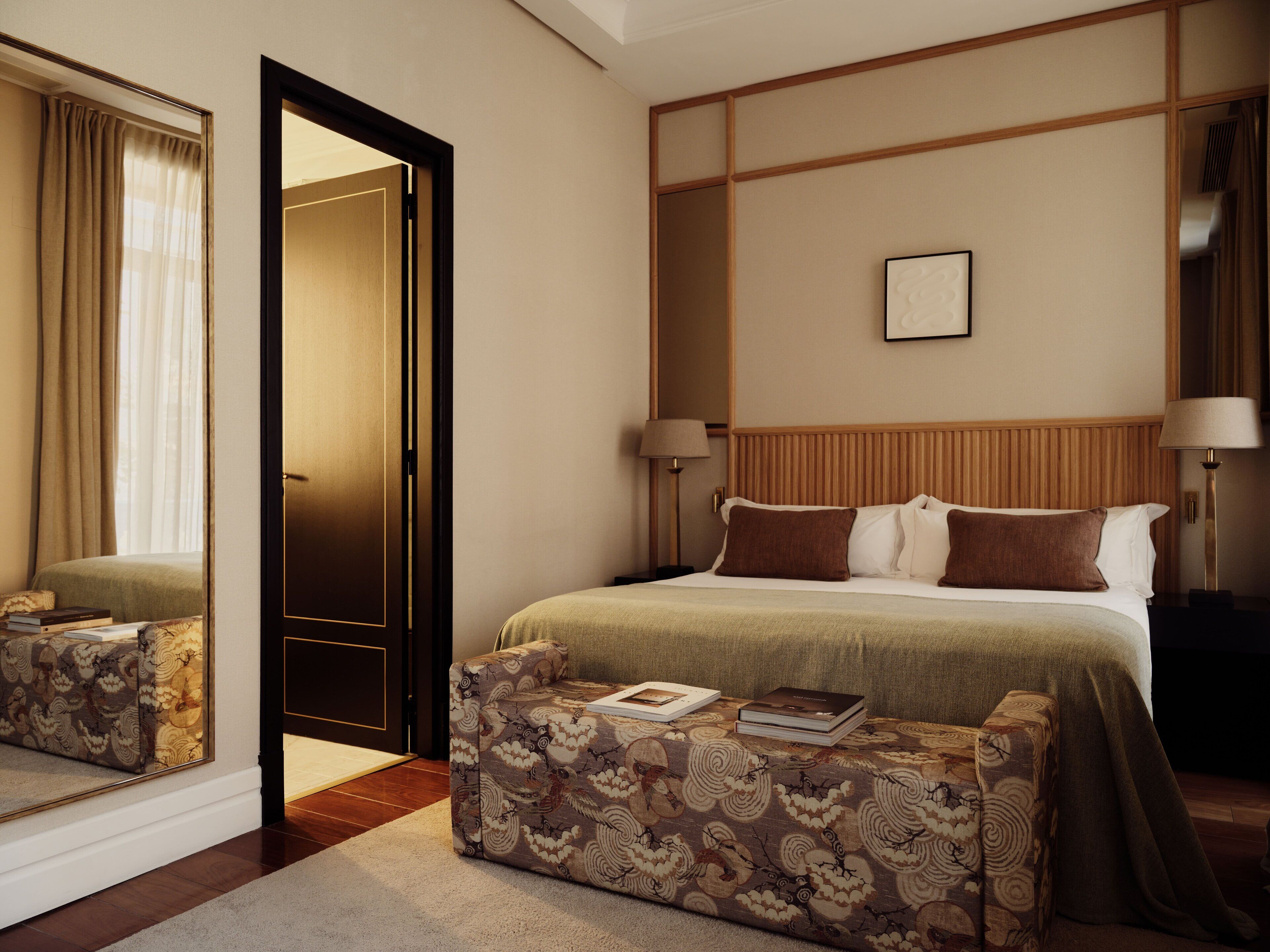 Quarto de hotel com cama, abajures, mesa de cabeceira com um livro, sofá e espelho.