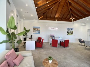 Um interior moderno com teto alto, balcão de recepção, cadeiras vermelhas, poltrona cinza e uma grande planta em vaso.