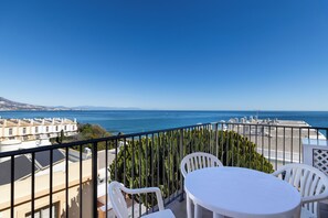 Varanda com mesa e cadeiras brancas, vista para a costa com edifícios e o mar.
