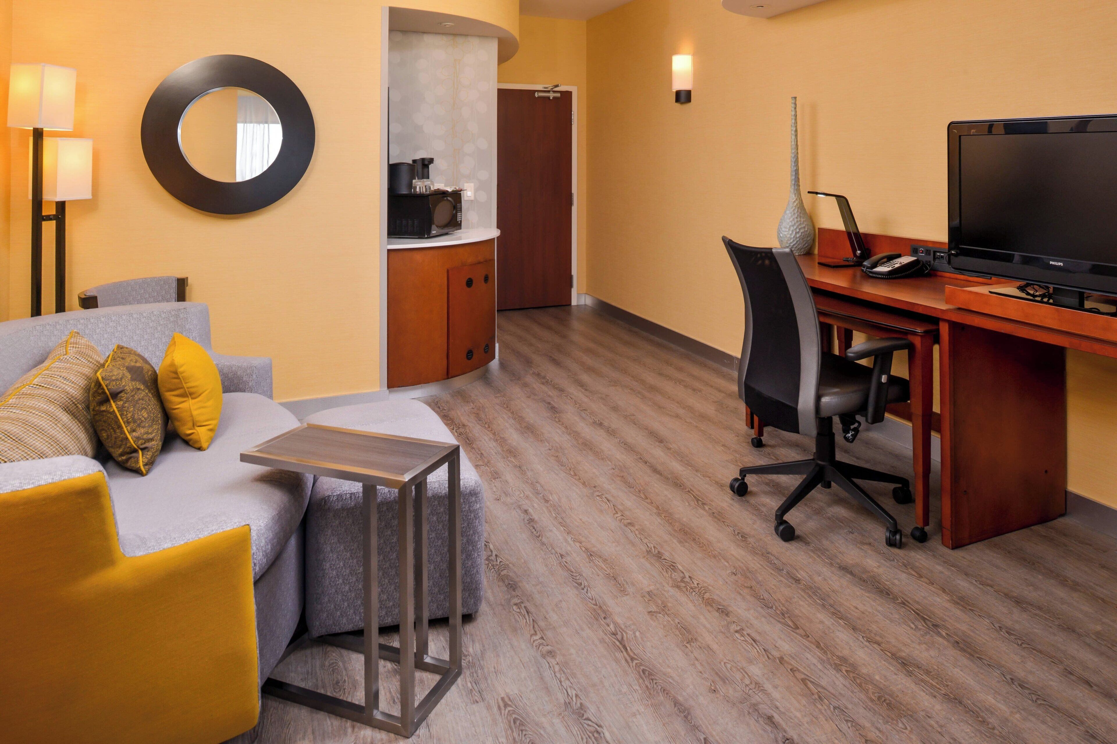 Quarto de hotel com sofá, escrivaninha com computador, luminária, espelho e máquina de café.