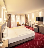 Quarto de hotel com uma cama grande, duas cadeiras, uma escrivaninha, televisão e uma janela com cortinas.