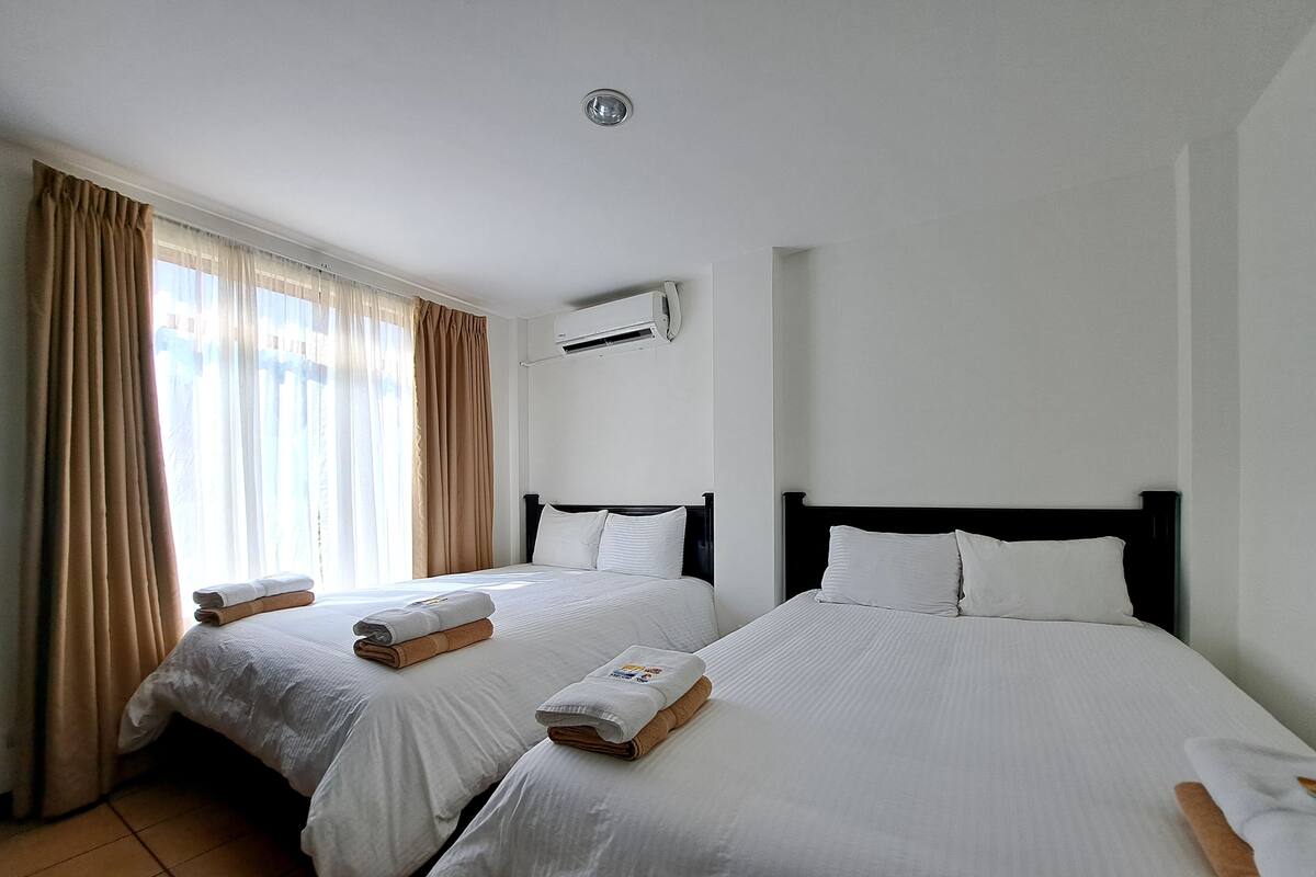 Quarto de hotel com duas camas, cortinas bege, uma luminária no teto e um ar condicionado.
