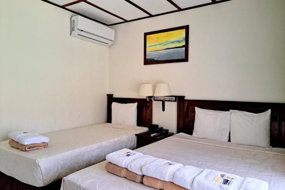 Quarto de hotel com duas camas, uma mesa de cabeceira, um abajur, ar condicionado e um quadro na parede.