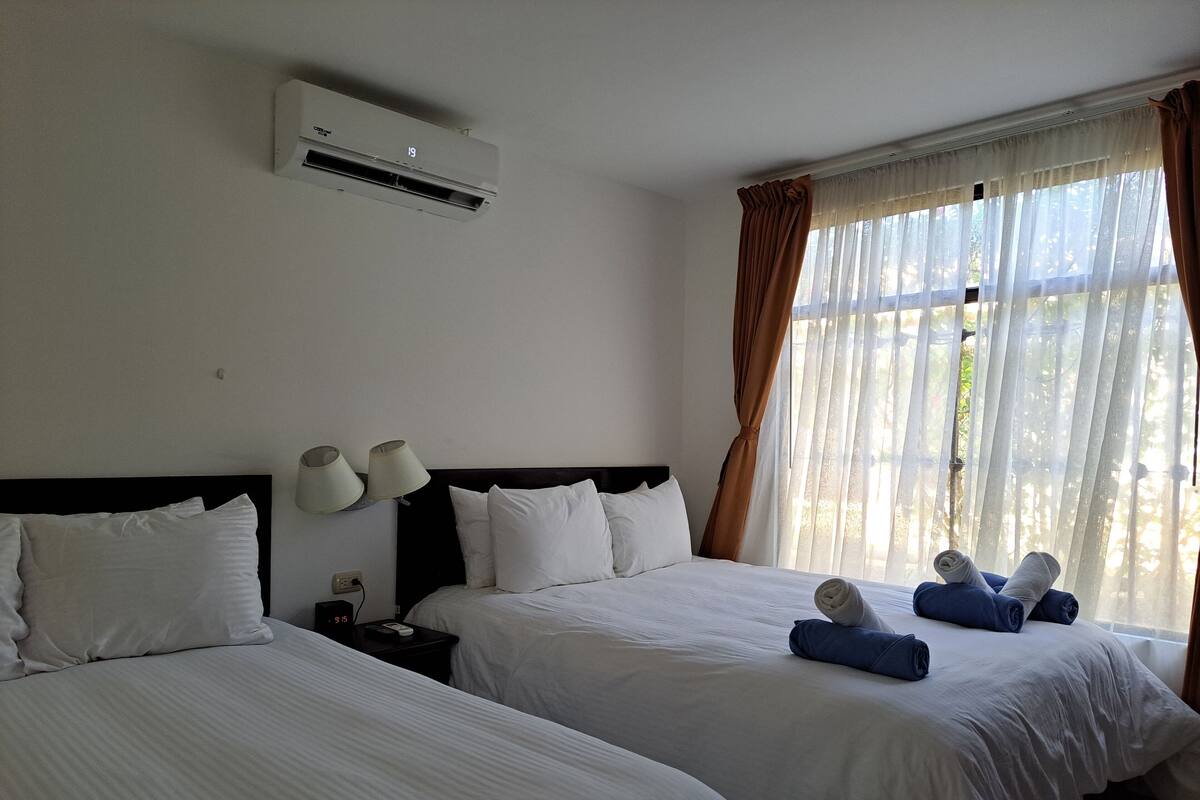 Quarto de hotel com duas camas, ar condicionado, mesa de cabeceira com abajur e janela com cortinas.