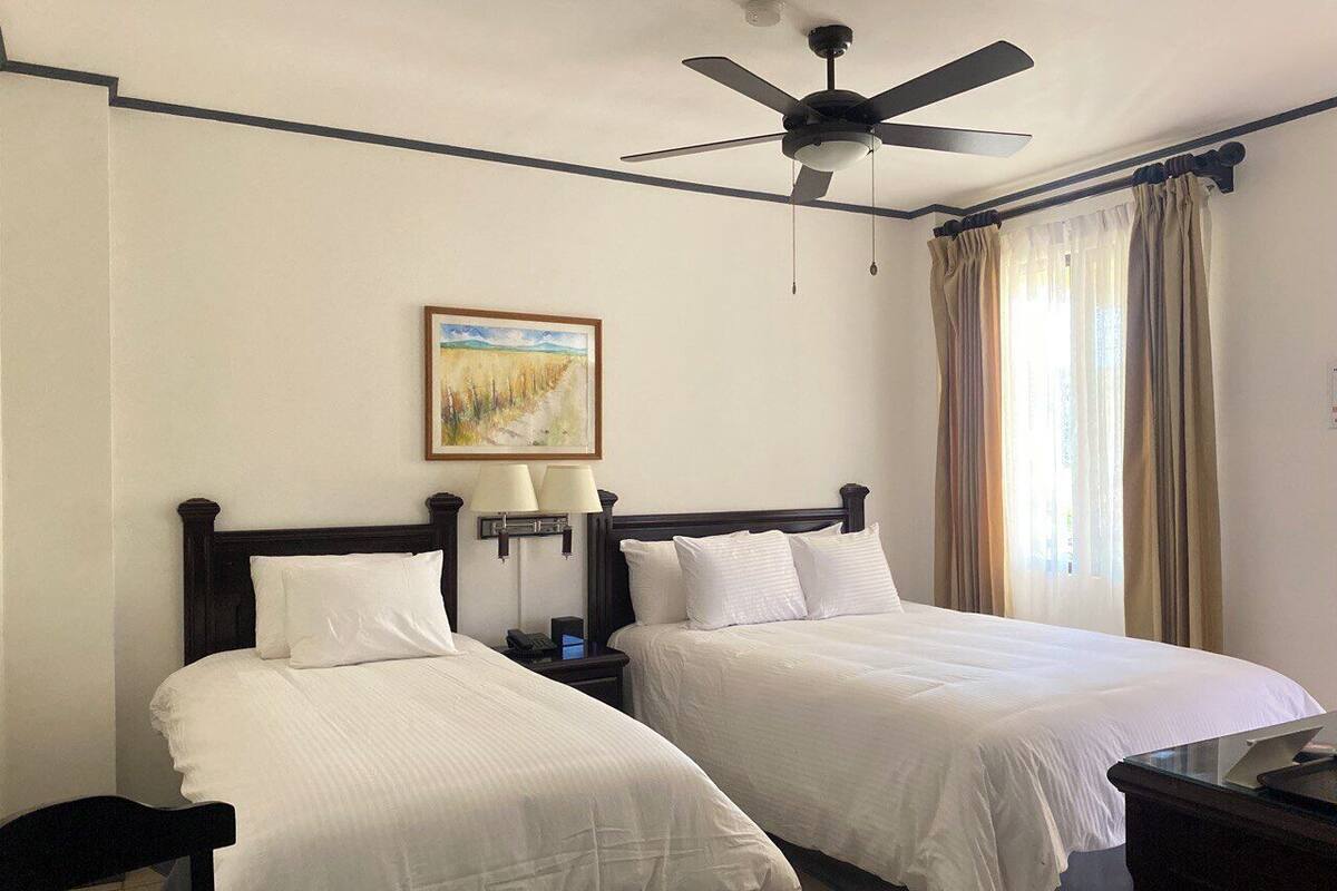 Quarto de hotel com duas camas de solteiro, um ventilador de teto, um quadro na parede e uma janela com cortinas.