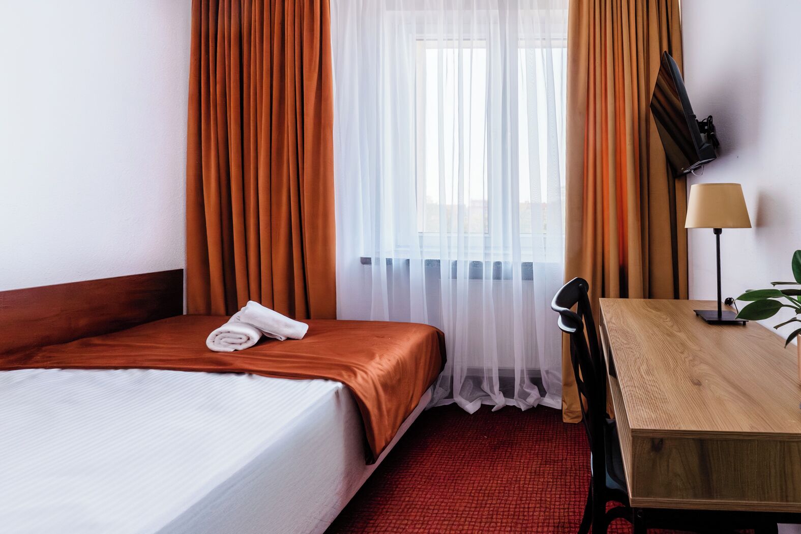 Quarto de hotel com cama, escrivaninha, cadeira, luminária e janela com cortinas.