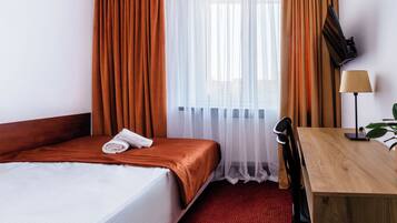 Quarto de hotel com cama, escrivaninha, cadeira, luminária e janela com cortinas.