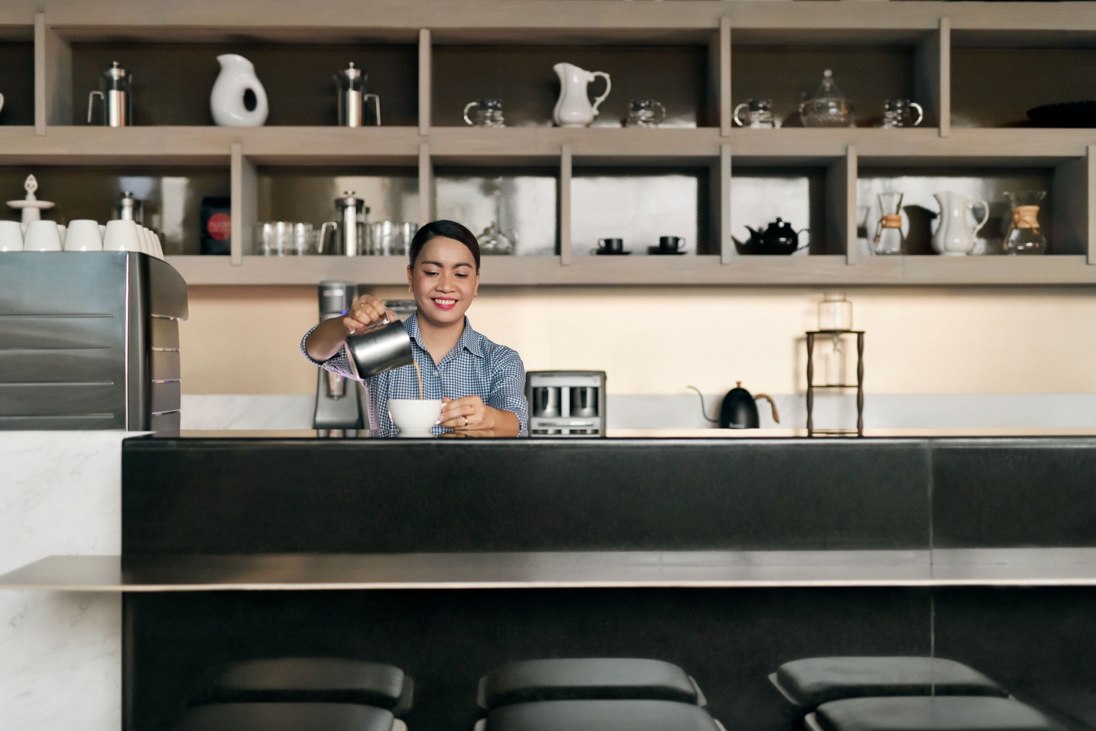 Um barista prepara uma bebida em um balcão, com uma máquina de café e diversos copos e canecas expostos.