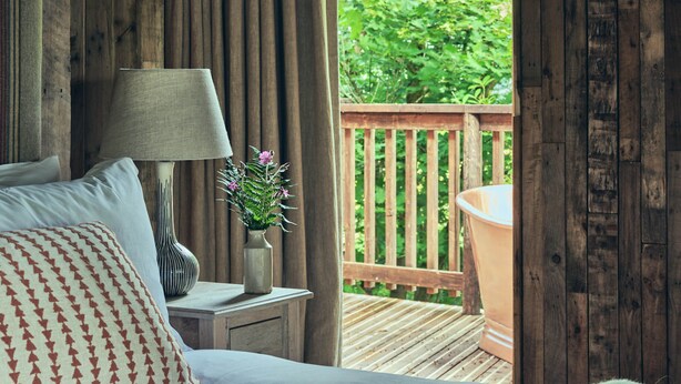Quarto de madeira com uma cama, mesa de cabeceira, abajur e vista para um deck de madeira e vegetação lá fora.