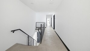 Um corredor comprido e estreito com paredes brancas, chão de concreto e escada de metal preta com corrimão.