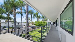 Uma varanda com guarda-corrimão de metal, chão de concreto e vista para um estacionamento com palmeiras e um edifício ao fundo.