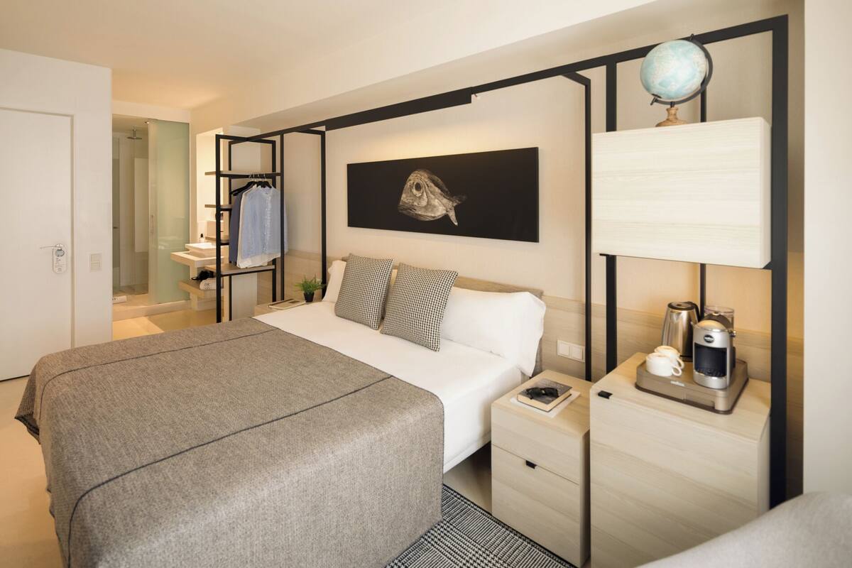Quarto de hotel moderno com uma cama, uma mesinha de cabeceira com abajur, um globo e uma cafeteira pequena.
