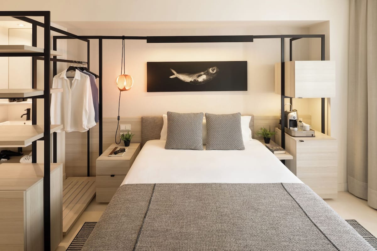 Quarto de hotel moderno com cama, luminária pendente, obra de arte na parede e mesa de cabeceira com abajur e máquina de café.