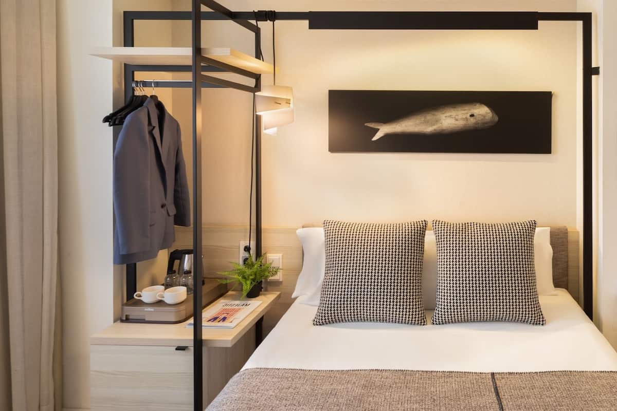 Quarto bem arrumado com cama, duas almofadas xadrez, prateleira fixada na parede com um casaco, quadro de um peixe e uma pequena planta.