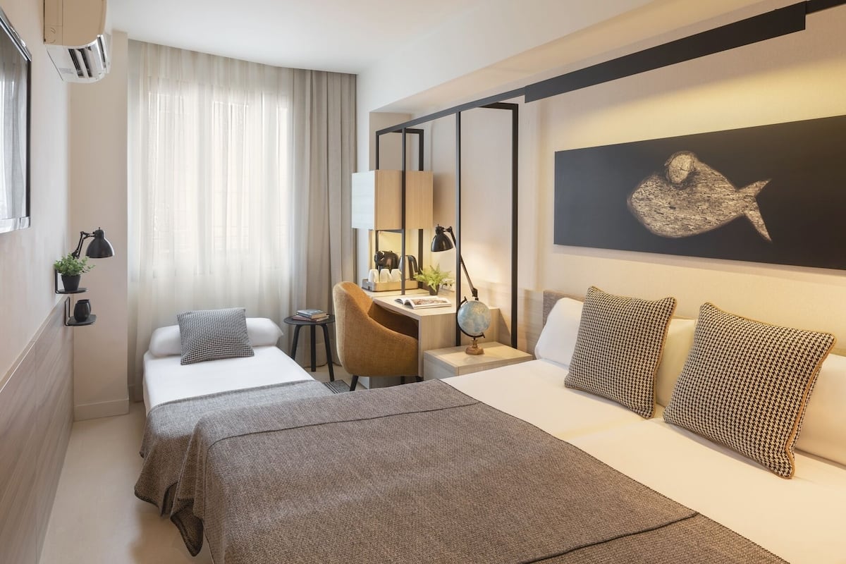 Quarto de hotel com duas camas, uma escrivaninha, uma cadeira, um globo e um quadro na parede.