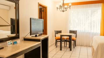 Quarto de hotel com cama, escrivaninha com computador, cadeira, mesa, lustre e janela com cortinas.
