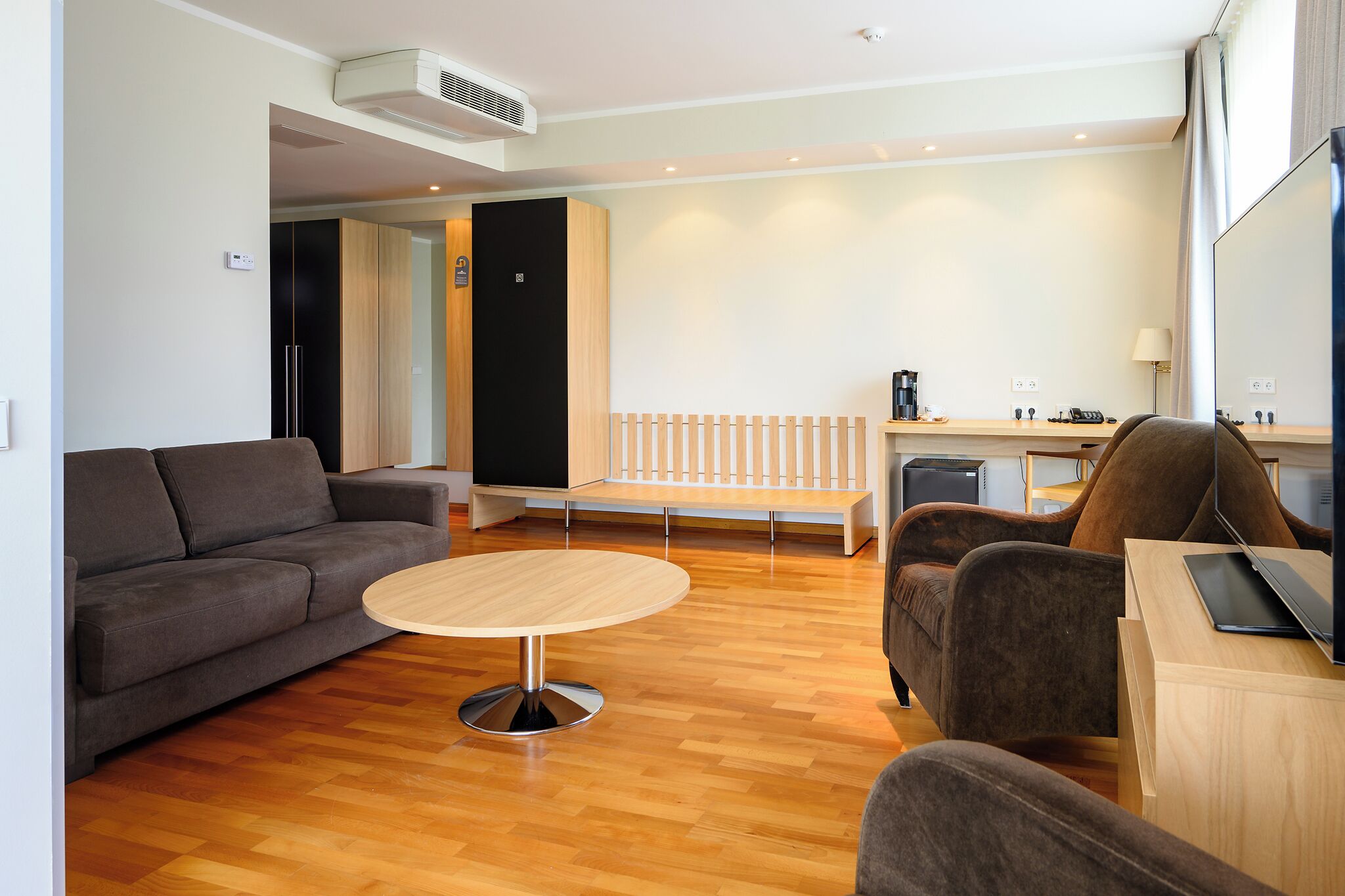 Uma sala de estar moderna com sofá, mesa redonda, poltrona marrom, banco de madeira, escrivaninha com laptop e televisão.