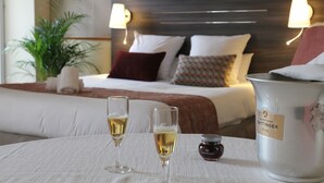 Quarto de hotel com cama bem arrumada, duas luminárias de cabeceira, uma garrafa de champanhe e duas taças de champanhe sobre uma mesa.
