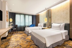 Quarto de hotel com duas camas, uma escrivaninha, uma cadeira e uma janela grande com cortinas.