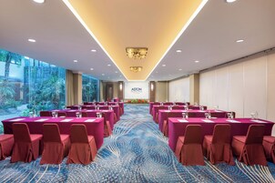 Sala de conferências com mesas, cadeiras, um ecrã grande e vista para o exterior arborizado.