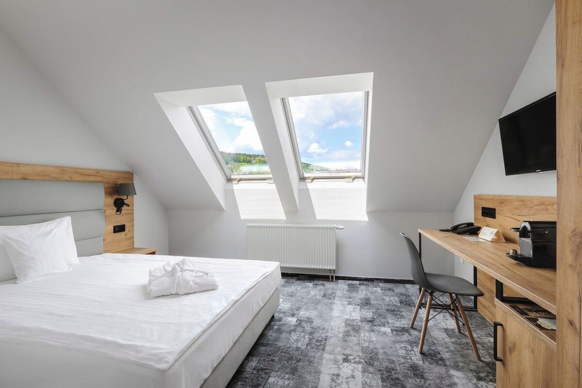 Quarto de hotel com cama, escrivaninha, cadeira, televisão e claraboias.