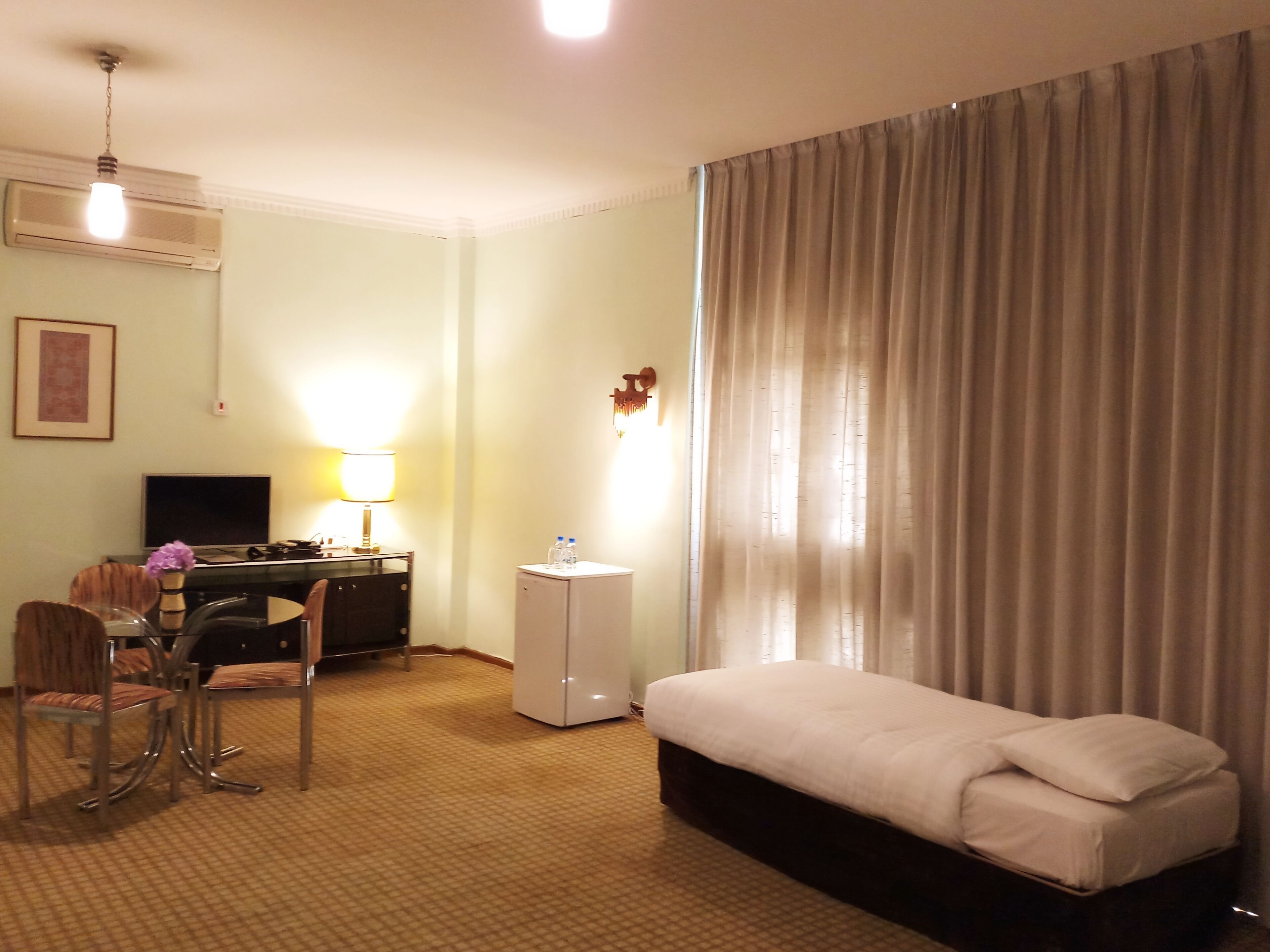 Quarto de hotel com cama, escrivaninha com cadeira, luminária, minibar e televisão.