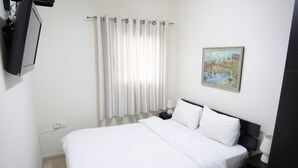 Quarto de hotel com uma cama, mesa de cabeceira, televisão fixada na parede, janela com cortinas e um quadro na parede.