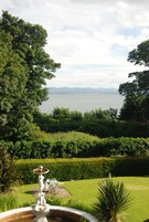 Um jardim com uma fonte, sebes bem aparadas e vista para um corpo de água e montanhas ao longe.