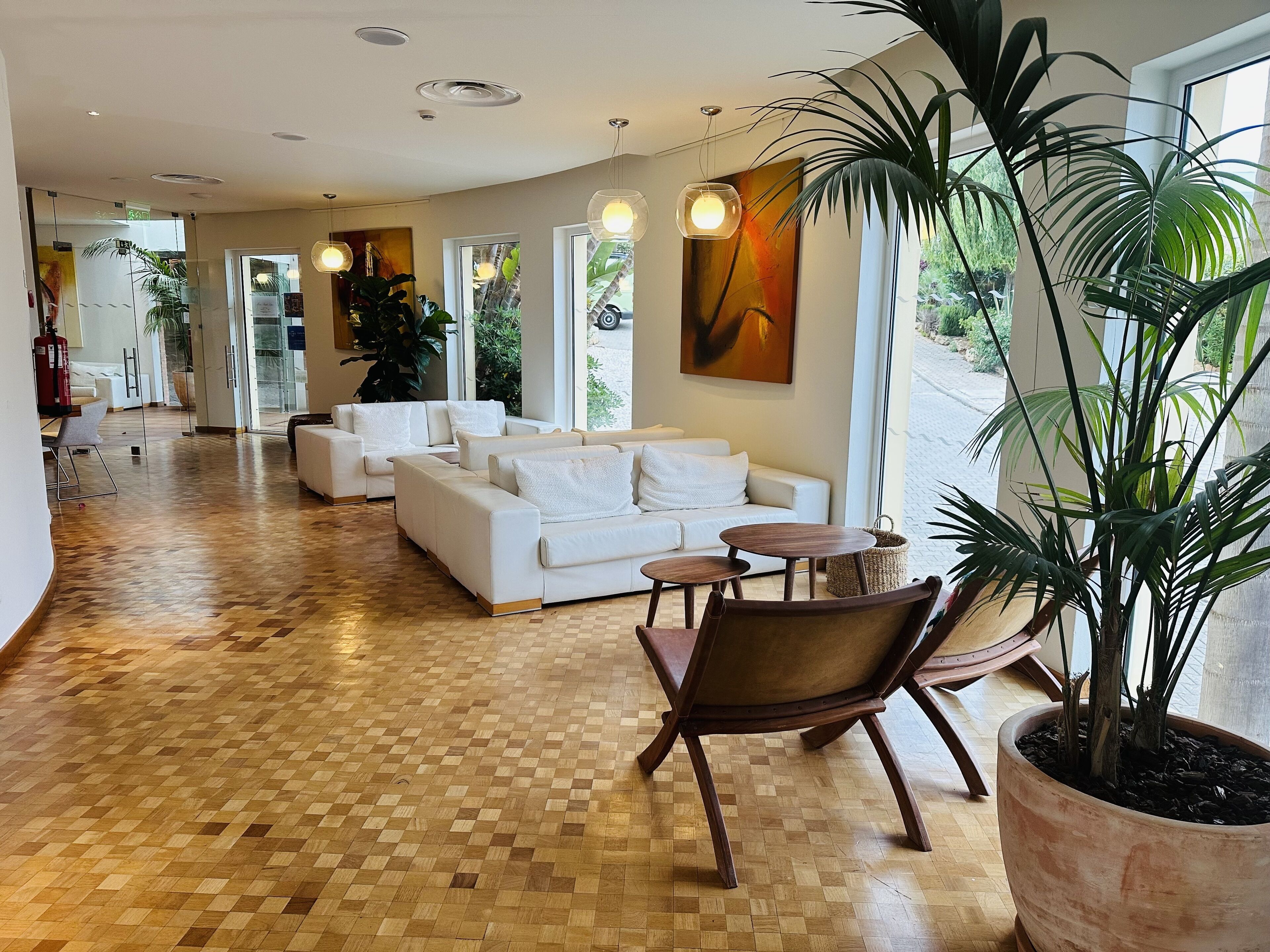 Um hall amplo com piso de madeira quadriculado, sofás brancos, uma cadeira de madeira e uma grande planta em vaso.
