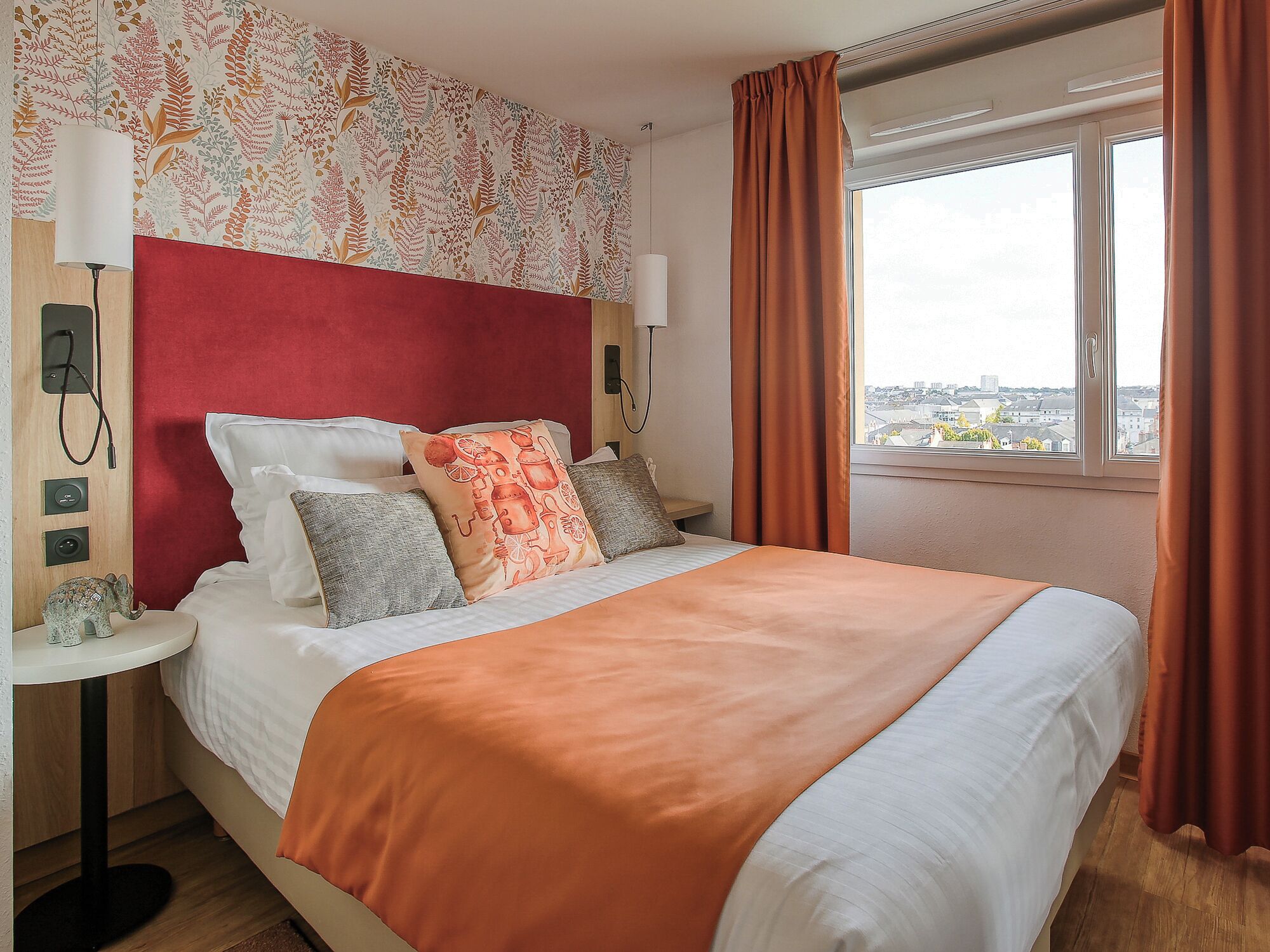 Quarto de hotel com uma cama grande, cabeceira vermelha, papel de parede estampado, vista da cidade pela janela e mesa de cabeceira com abajur.