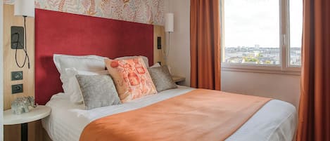 Quarto de hotel com uma cama grande, cabeceira vermelha, papel de parede estampado, vista da cidade pela janela e mesa de cabeceira com abajur.