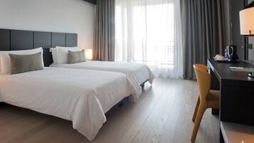 Quarto de hotel com duas camas, uma escrivaninha com cadeira e uma janela grande com cortinas.