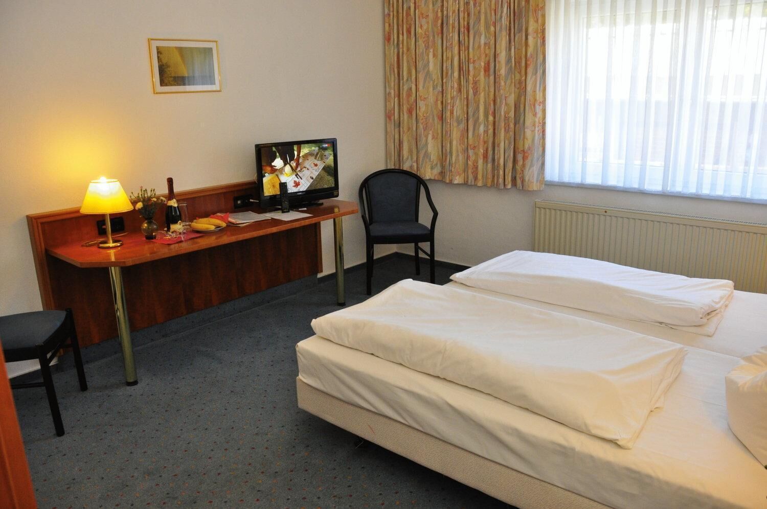 Quarto de hotel com duas camas, uma escrivaninha com luminária, uma cadeira, uma televisão e uma janela com cortinas.