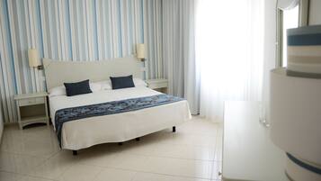 Cama bem arrumada com colcha branca e azul estampada, duas almofadas escuras e cabeceira combinando. Há duas mesas de cabeceira com abajures, uma janela grande com cortinas leves e uma parede branca com listras verticais.